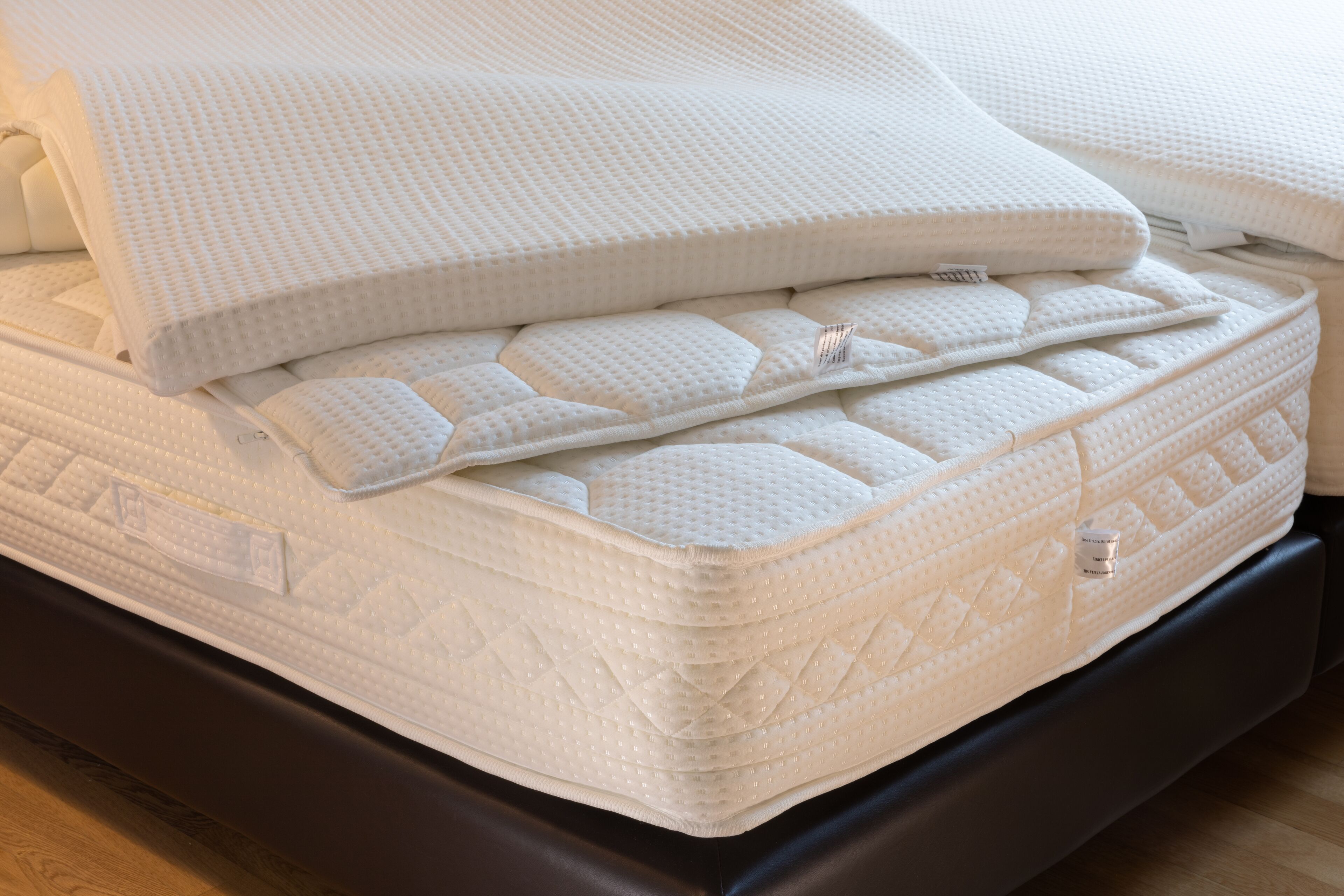 Uma cama bem arrumada com colchão e almofadas brancas, cabeceira escura e piso de madeira.