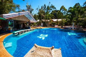 Piscina com deck revestido, área de estar coberta e árvores ao redor.