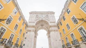 Um arco grande e ornamentado com detalhes arquitetônicos clássicos, ladeado por edifícios amarelos com várias janelas e varandas.