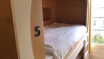 Uma cama beliche com o número '5' na parede, cabeceira de madeira e uma janela com vista para edifícios.