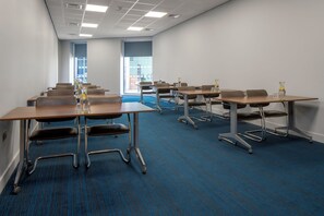 Sala de conferência com várias mesas e cadeiras, carpete azul e vista para o exterior através de grandes janelas.