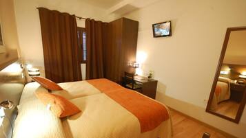 Quarto de hotel com cama de casal, escrivaninha, cadeira, mesa pequena com abajur, espelho e quadro na parede.
