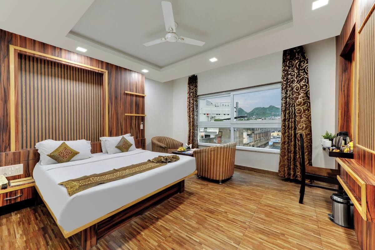 Quarto de hotel com uma cama grande, uma cadeira, uma mesinha, uma janela com vista e um ventilador de teto.
