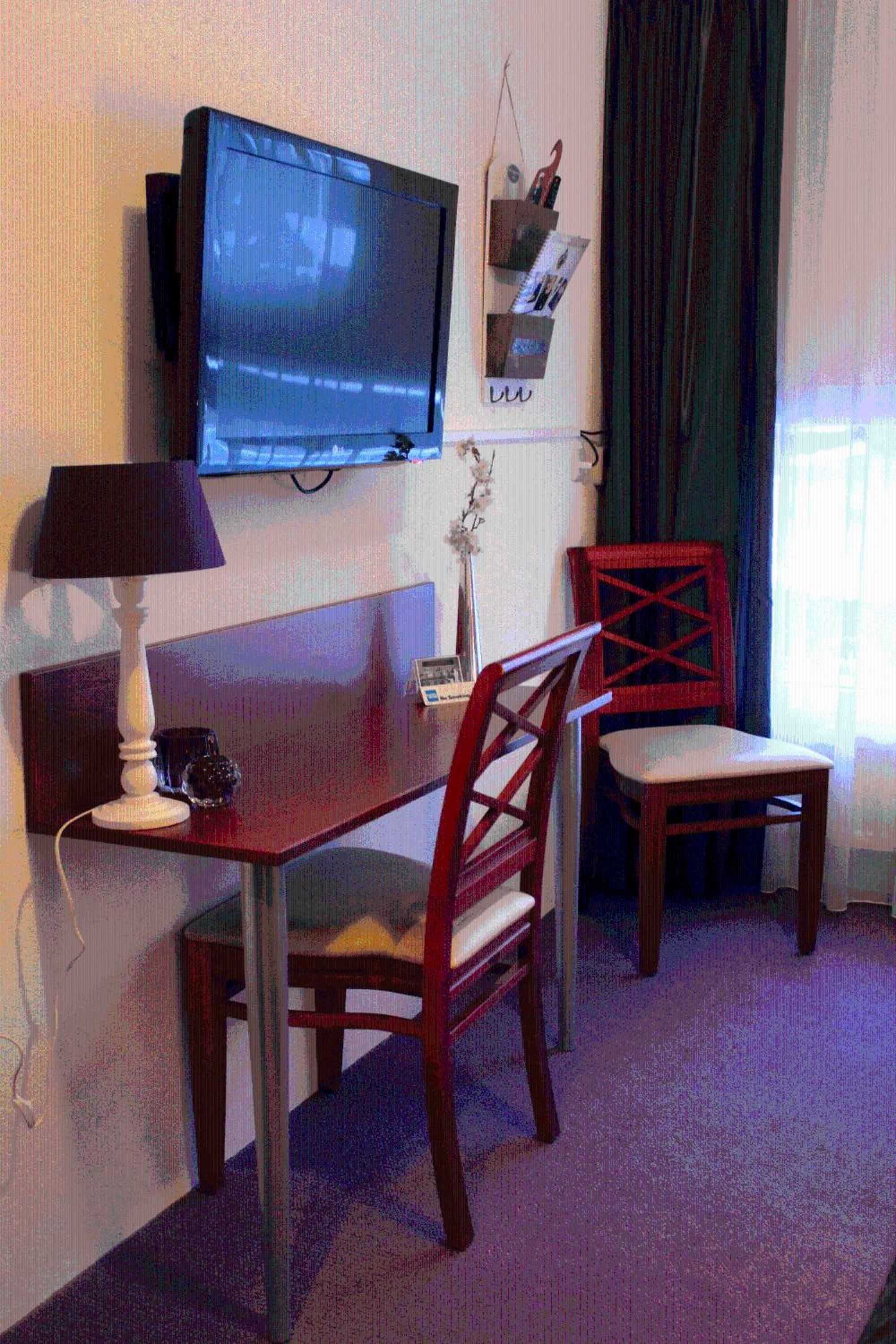 Quarto de hotel com televisão na parede, escrivaninha de madeira, duas cadeiras, uma lâmpada e uma pequena estante com revistas.