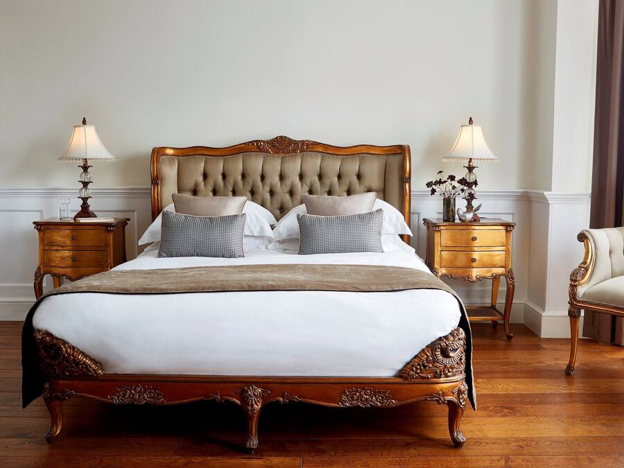 Uma cama grande e ricamente decorada com cabeceira estofada, acompanhada por duas mesas de cabeceira com abajures, em um quarto com piso de madeira e uma poltrona clássica.