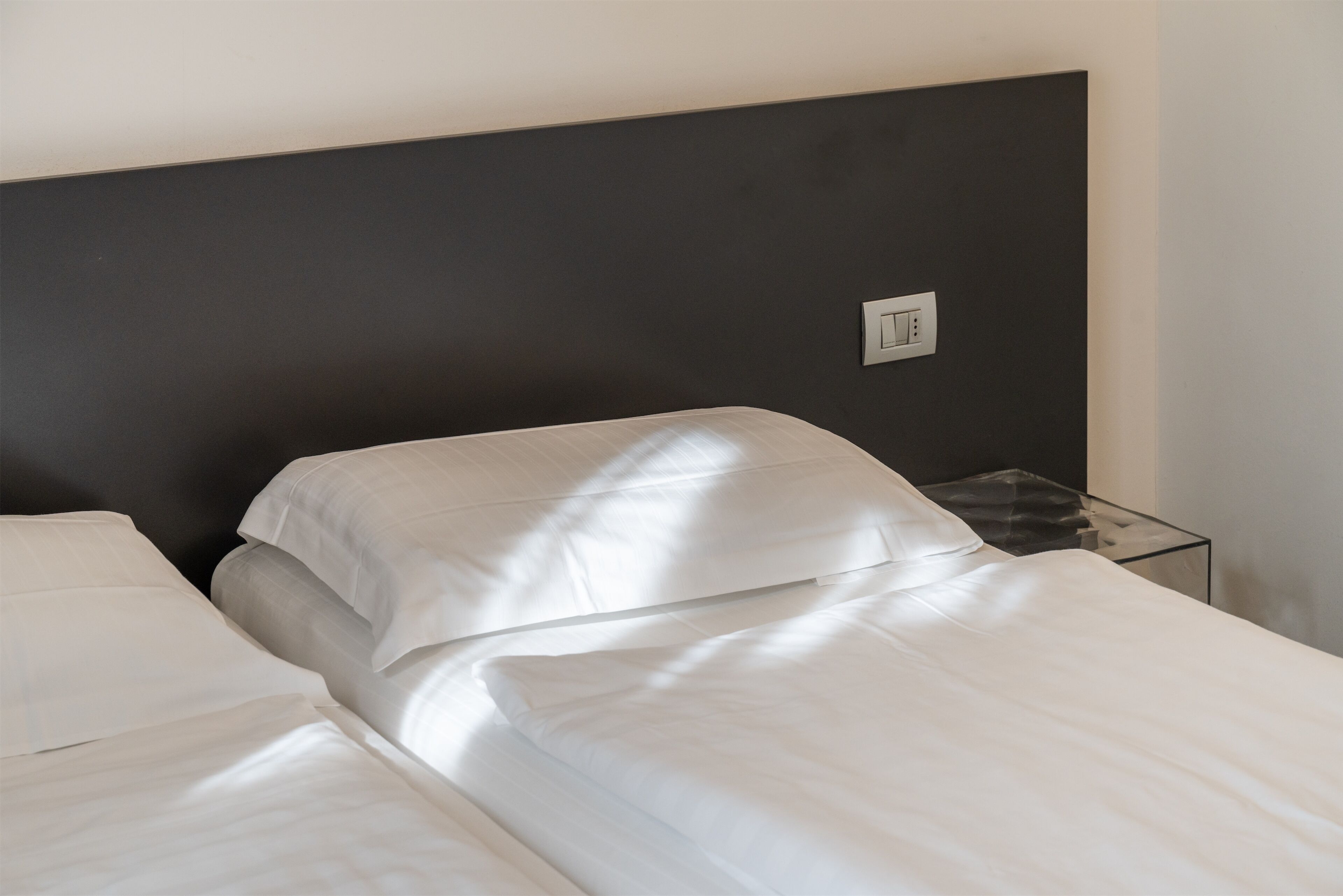 Uma cama de solteiro com roupa de cama branca em quarto com paredes escuras e uma tomada elétrica à vista.