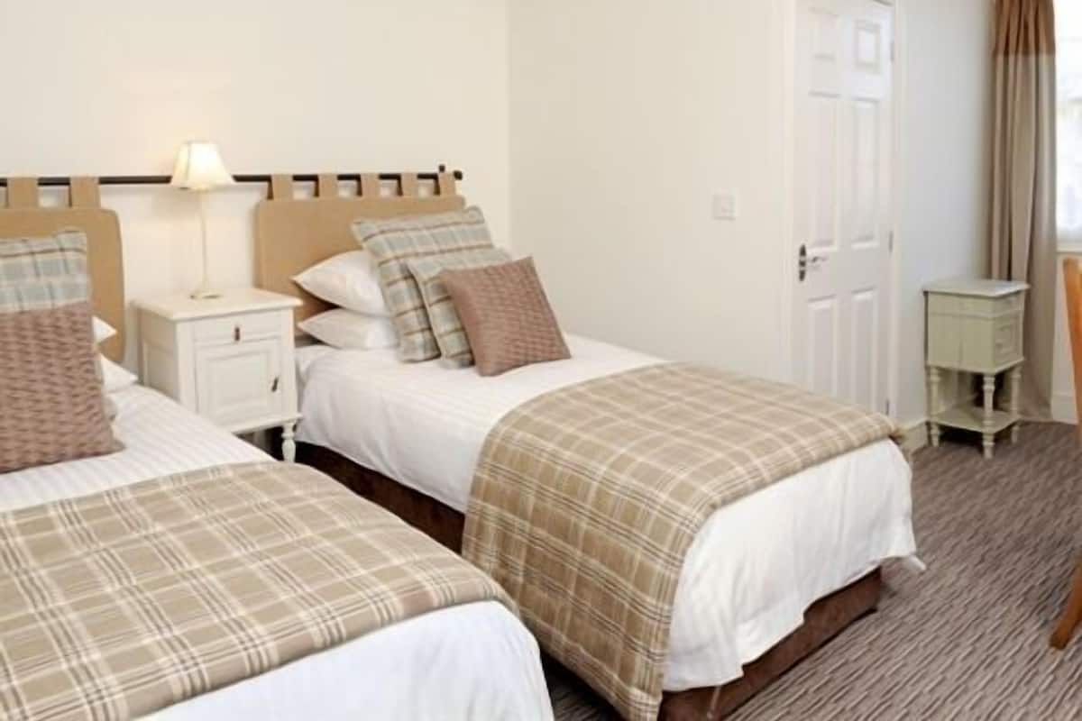 Quarto de hotel com duas camas, uma mesa de cabeceira, uma luminária e uma porta.