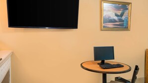 Quarto com televisão de tela plana na parede, uma mesa redonda pequena com computador e teclado, uma cadeira e um quadro pendurado acima da mesa.