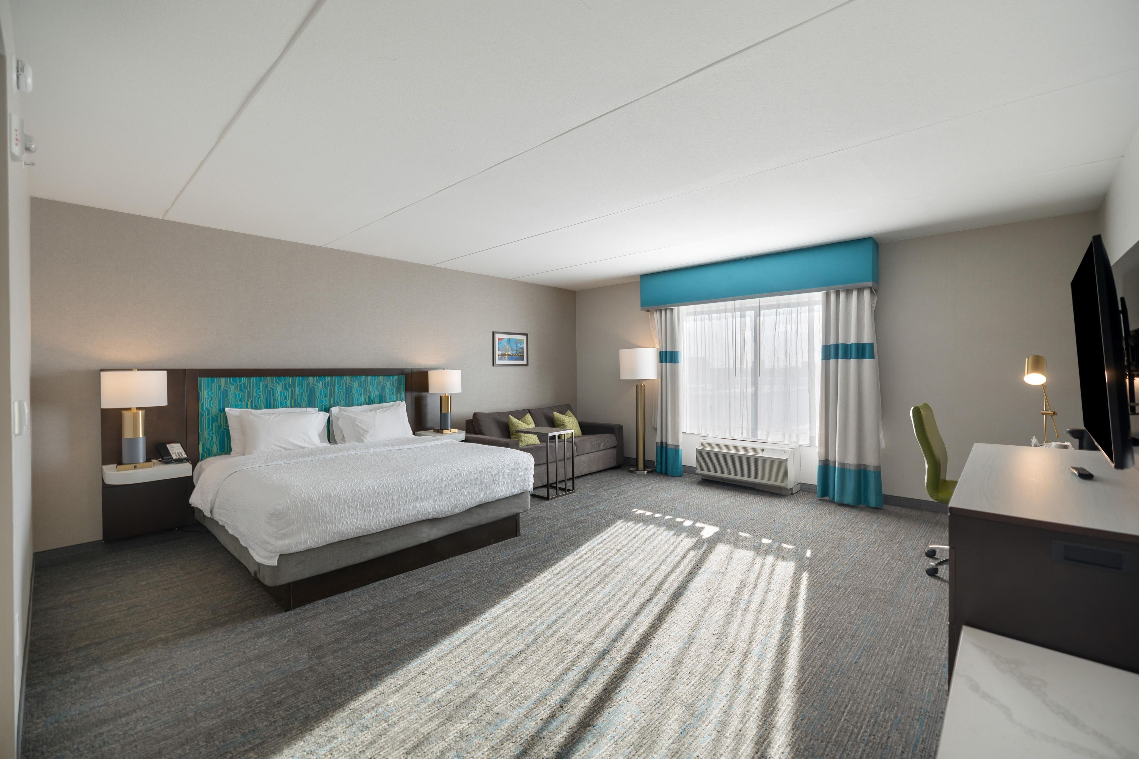 Quarto de hotel com cama grande, escrivaninha, cadeira, sofá, televisão e janela com cortinas.