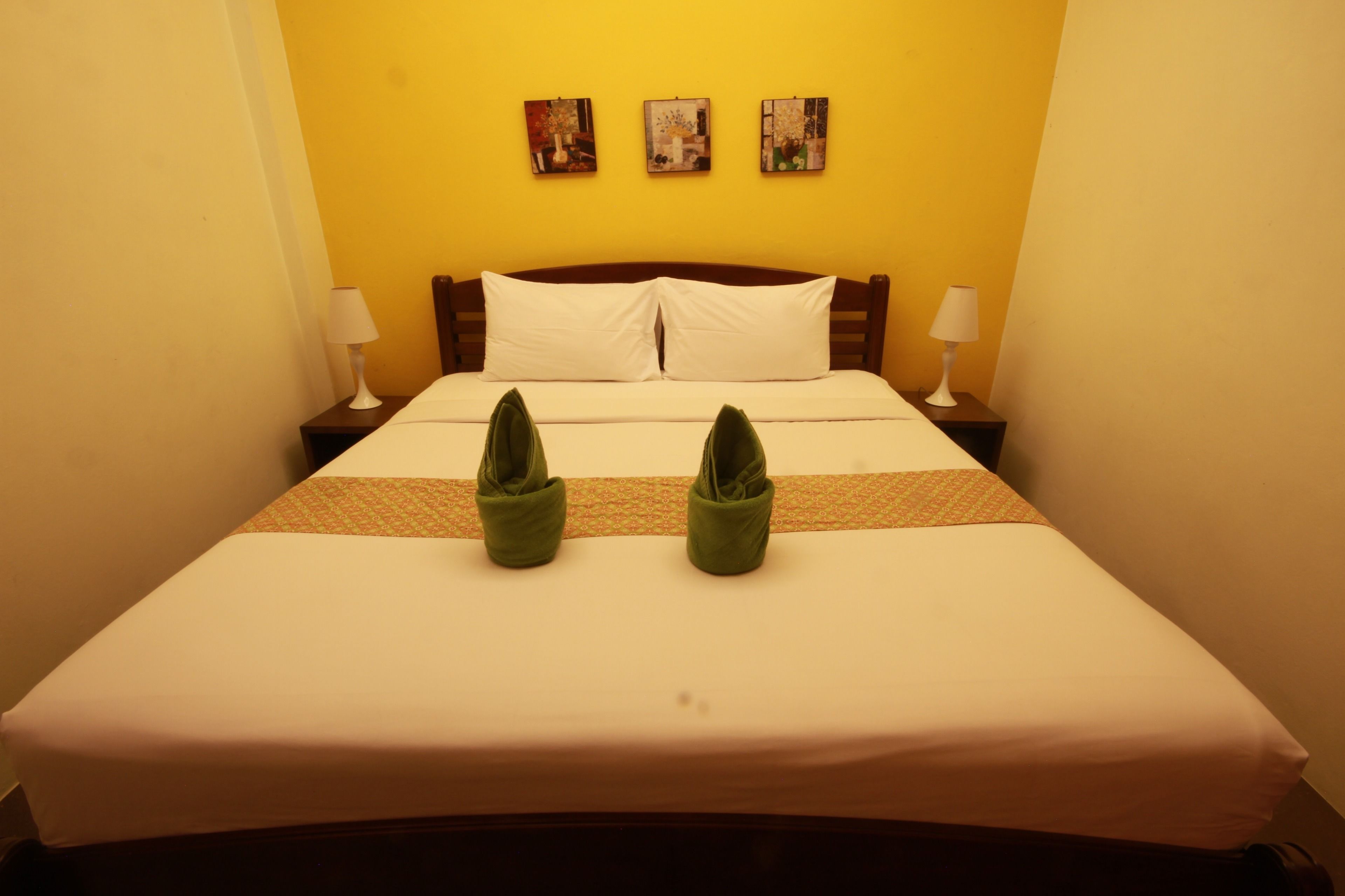 Cama bem arrumada com roupa de cama branca e duas toalhas verdes dobradas sobre a cama. Há duas mesas de cabeceira com abajures, e três quadros emoldurados na parede acima da cama.