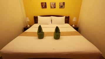 Cama bem arrumada com roupa de cama branca e duas toalhas verdes dobradas sobre a cama. Há duas mesas de cabeceira com abajures, e três quadros emoldurados na parede acima da cama.
