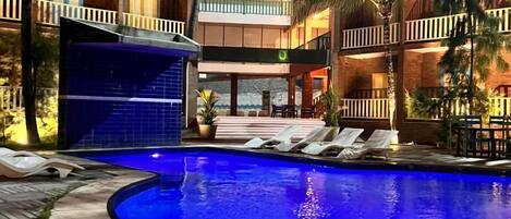 Uma piscina com espreguiçadeiras, palmeiras e um edifício com varandas.