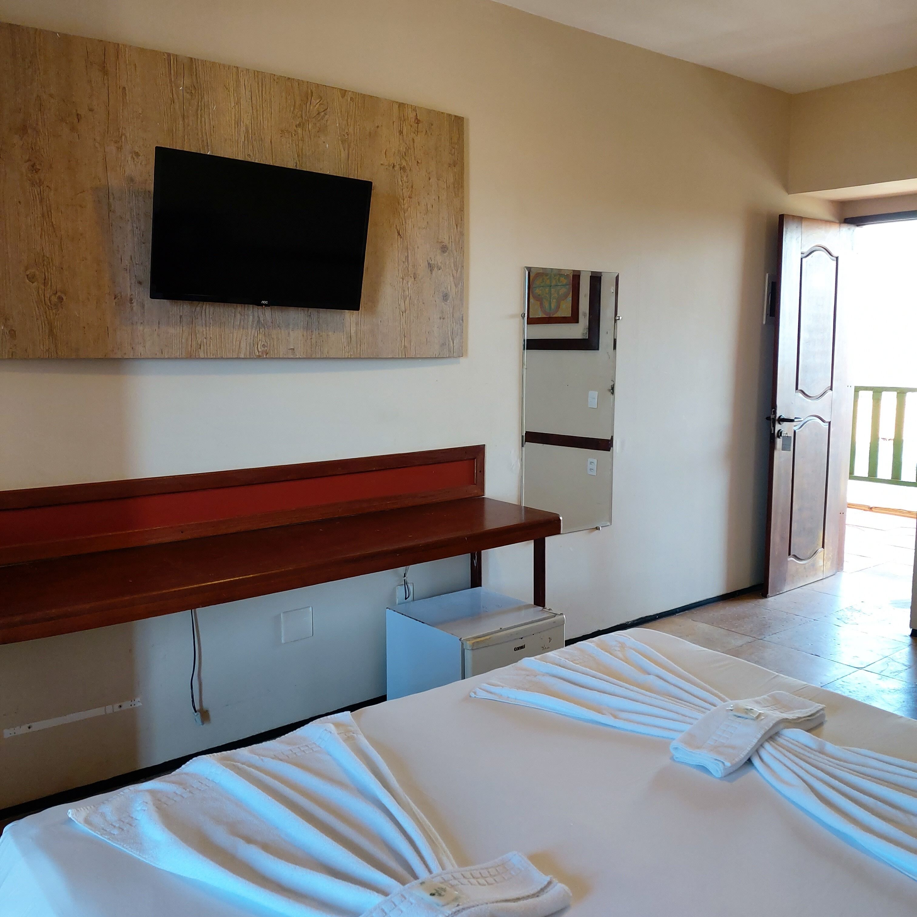 Quarto de hotel com cama, televisão fixada na parede, espelho e porta que dá para uma varanda.