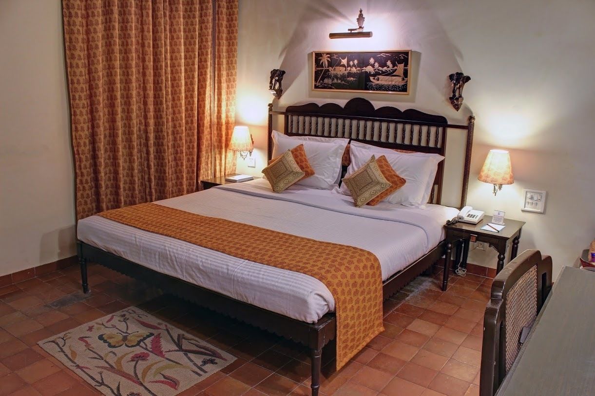 Uma cama bem arrumada com colcha estampada e almofadas, com duas mesas de cabeceira com luminárias, num quarto com chão de azulejos e um tapete decorativo.