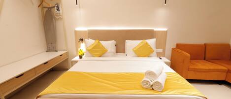 Quarto de hotel com cama, um banco, sofá laranja e mesa de cabeceira com luminária.