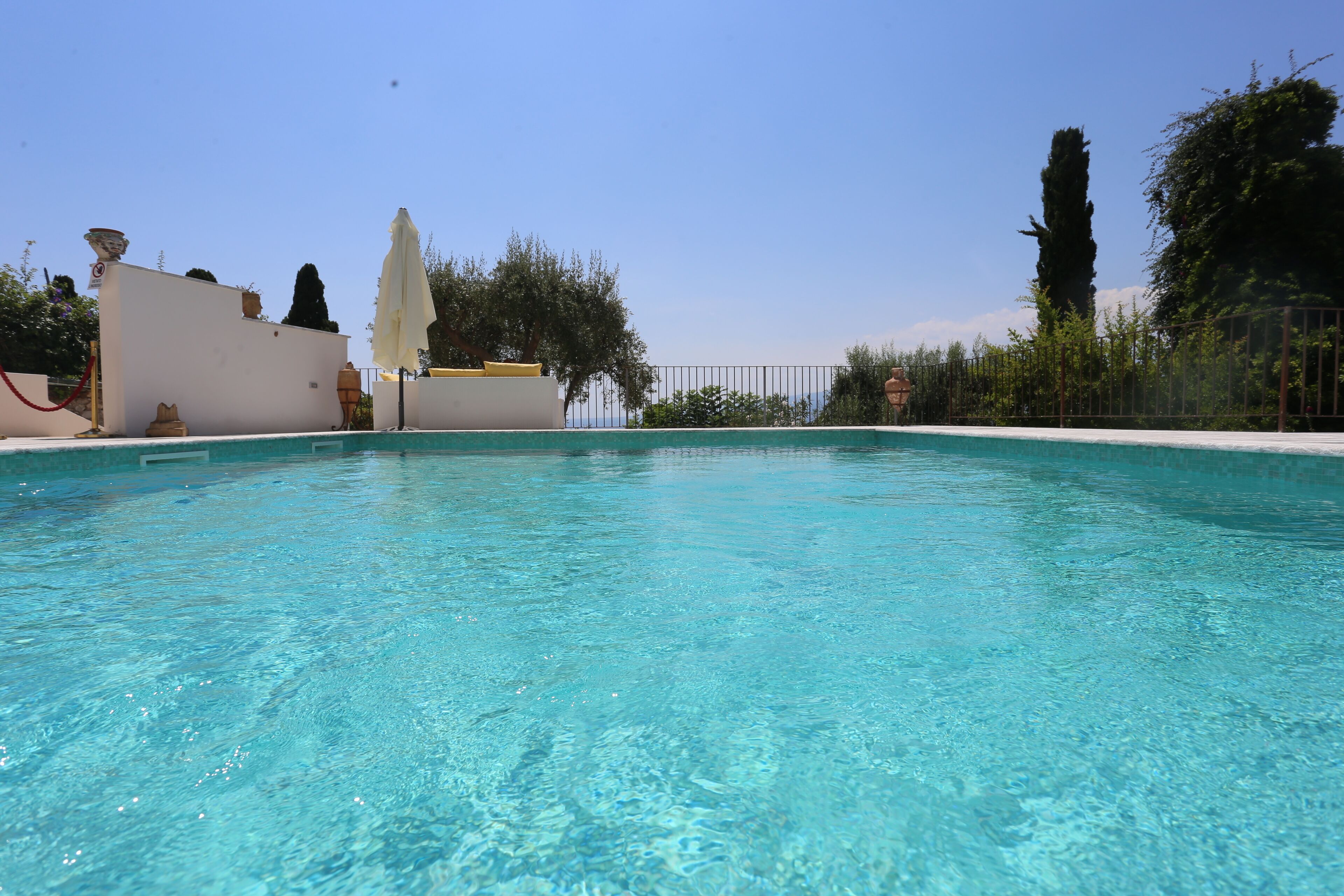 Piscina azul cristalina rodeada por um muro branco e vegetação.