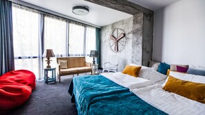 Quarto de hotel moderno com uma cama grande, um puff vermelho, um sofá e uma cadeira. Há uma janela grande com cortinas, um relógio de parede e uma luminária no teto.