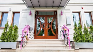 Um edifício com uma placa indicando "CITY SOLEI" e uma porta dupla de madeira. Duas bicicletas com suportes rosa estão estacionadas na frente.
