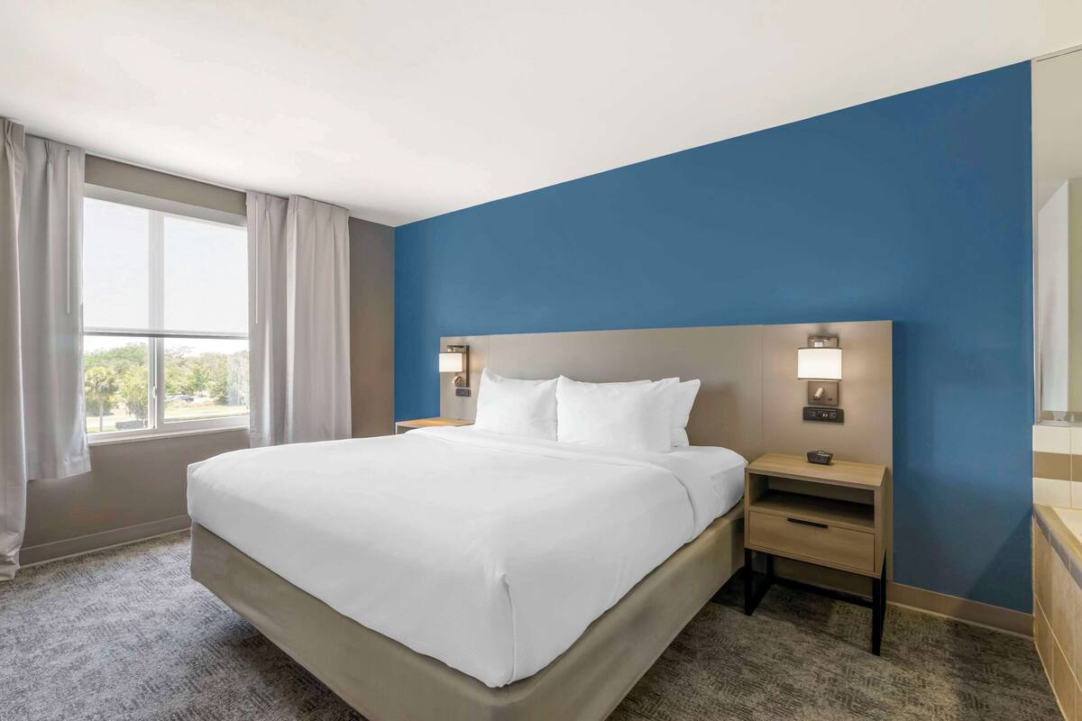 Quarto de hotel com uma cama grande, duas mesas de cabeceira com luminárias, uma janela com cortinas e uma parede azul.