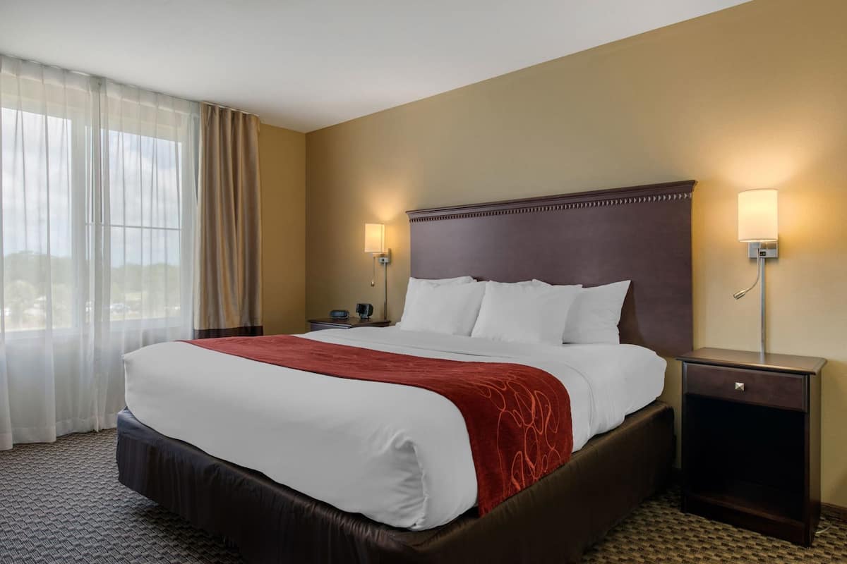 Quarto de hotel com cama grande, duas luminárias de cabeceira, mesa de apoio e vista pela janela.