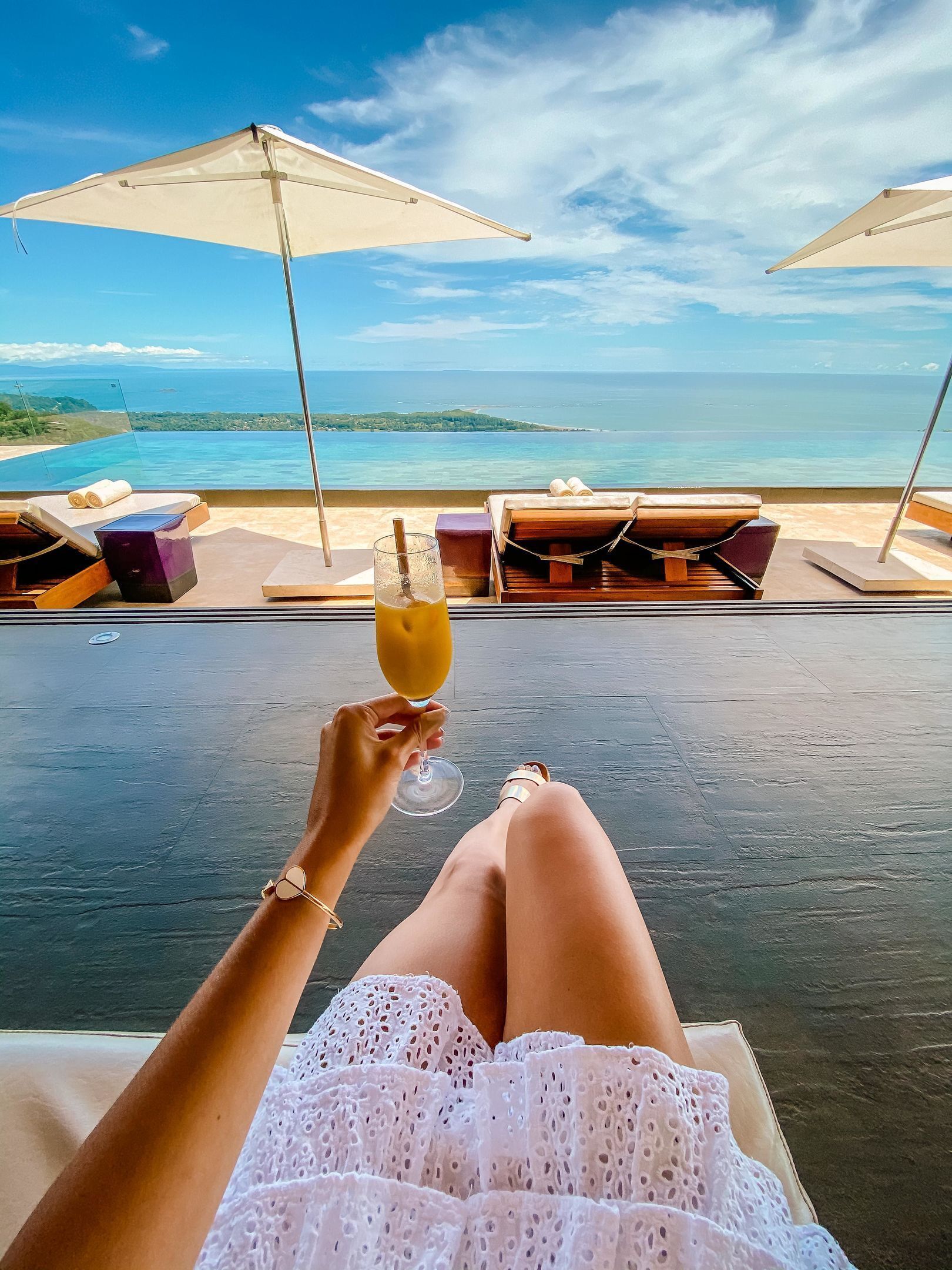 Uma pessoa relaxando em uma espreguiçadeira com uma bebida, com vista para uma praia com guarda-sóis e cadeiras de praia.
