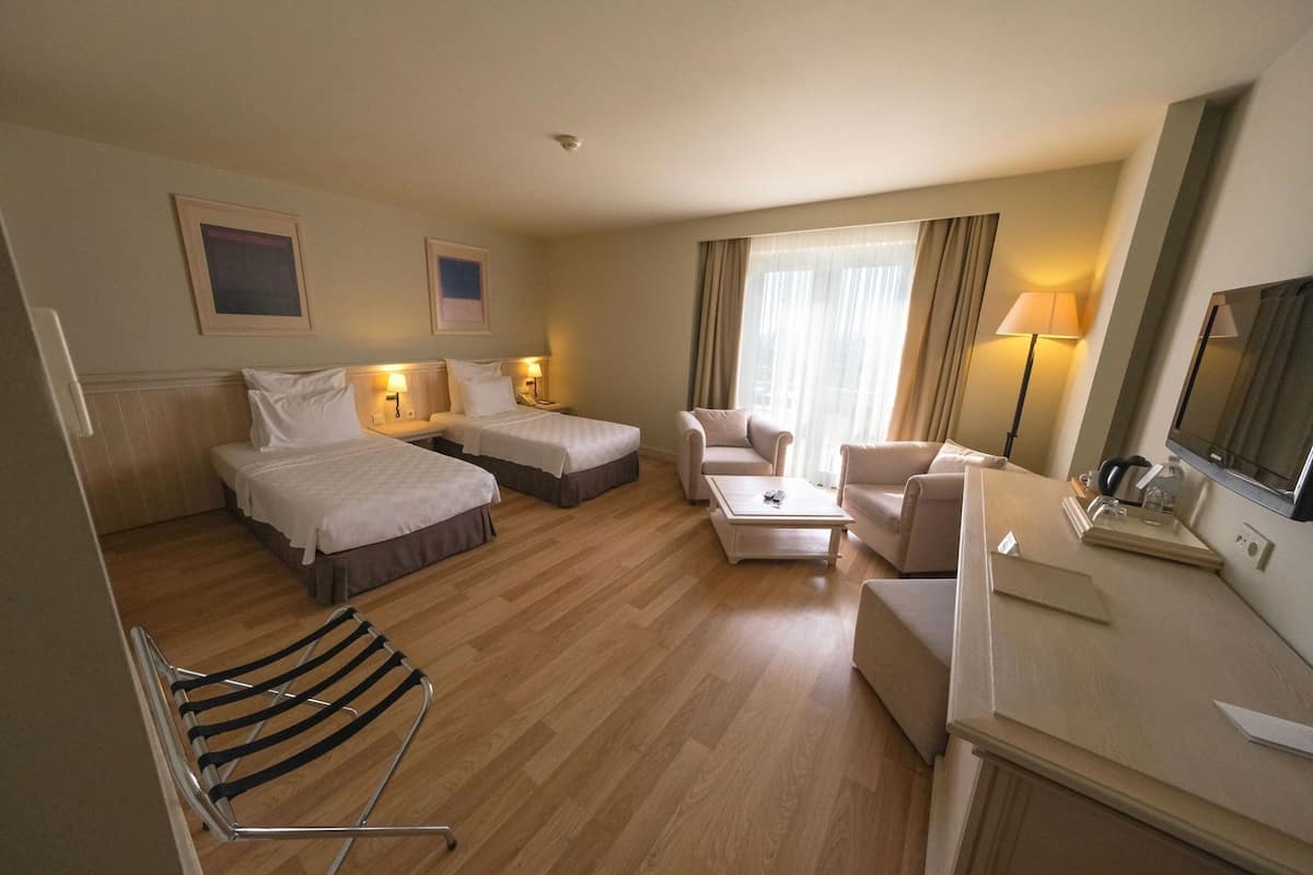 Quarto de hotel com duas camas, um sofá, uma poltrona, uma escrivaninha e uma televisão.