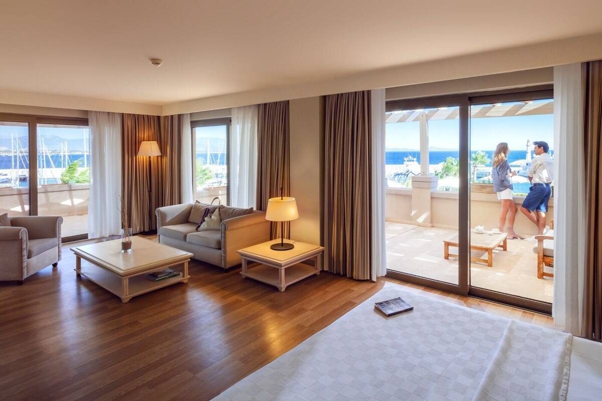 Quarto de hotel com uma cama grande, um sofá, uma mesa de centro e uma televisão. Há uma varanda com vista para o mar e um casal ao lado do corrimão.