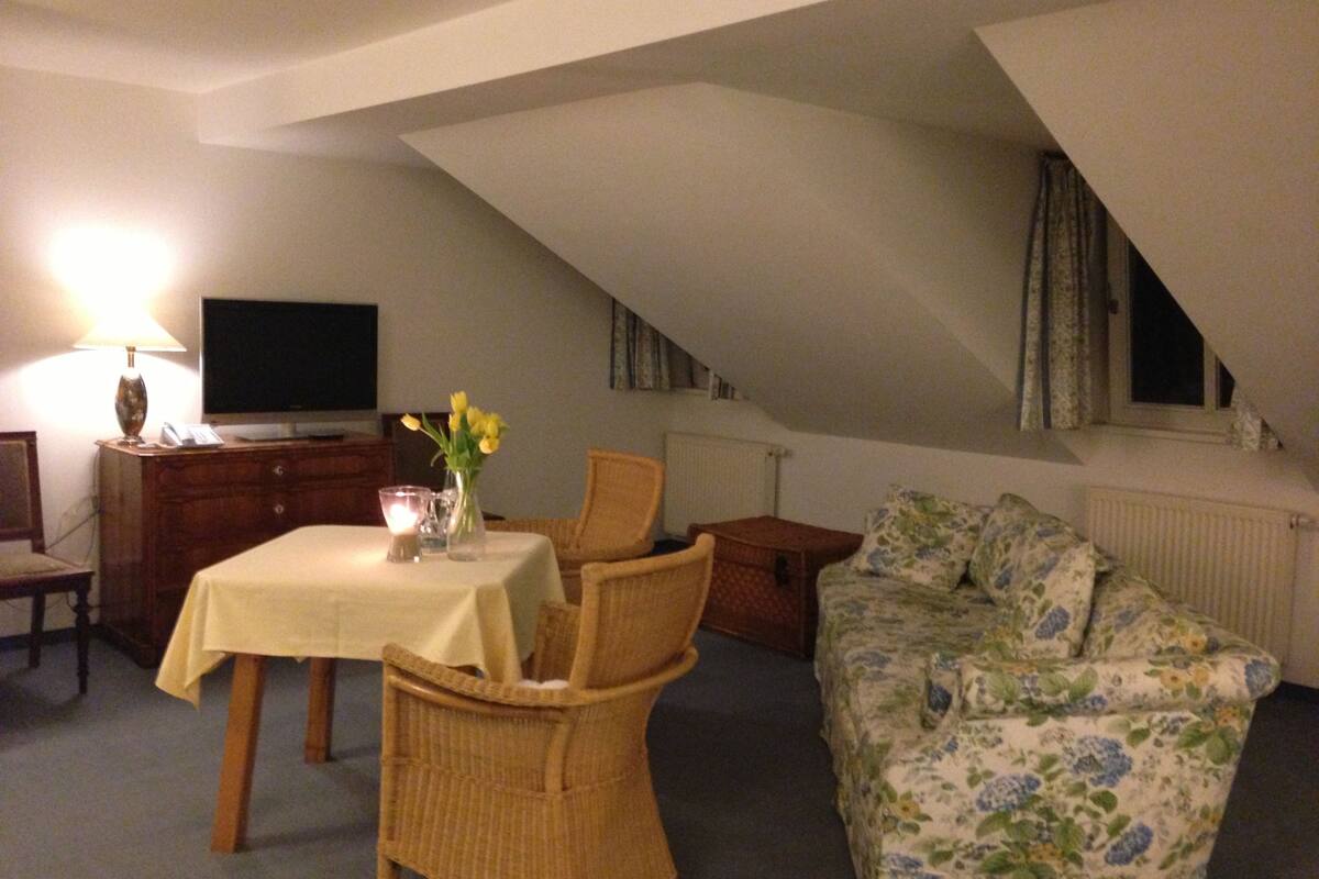 Um quarto de sótão com uma cama, uma mesa com um vaso de flores, uma cadeira, uma televisão e um aparador.