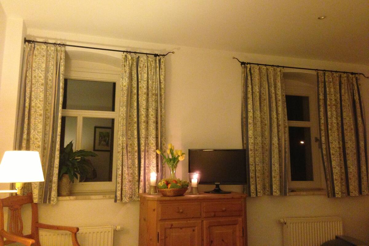 Quarto com duas janelas, um armário de madeira com televisão de tela plana, uma luminária e um vaso com flores.