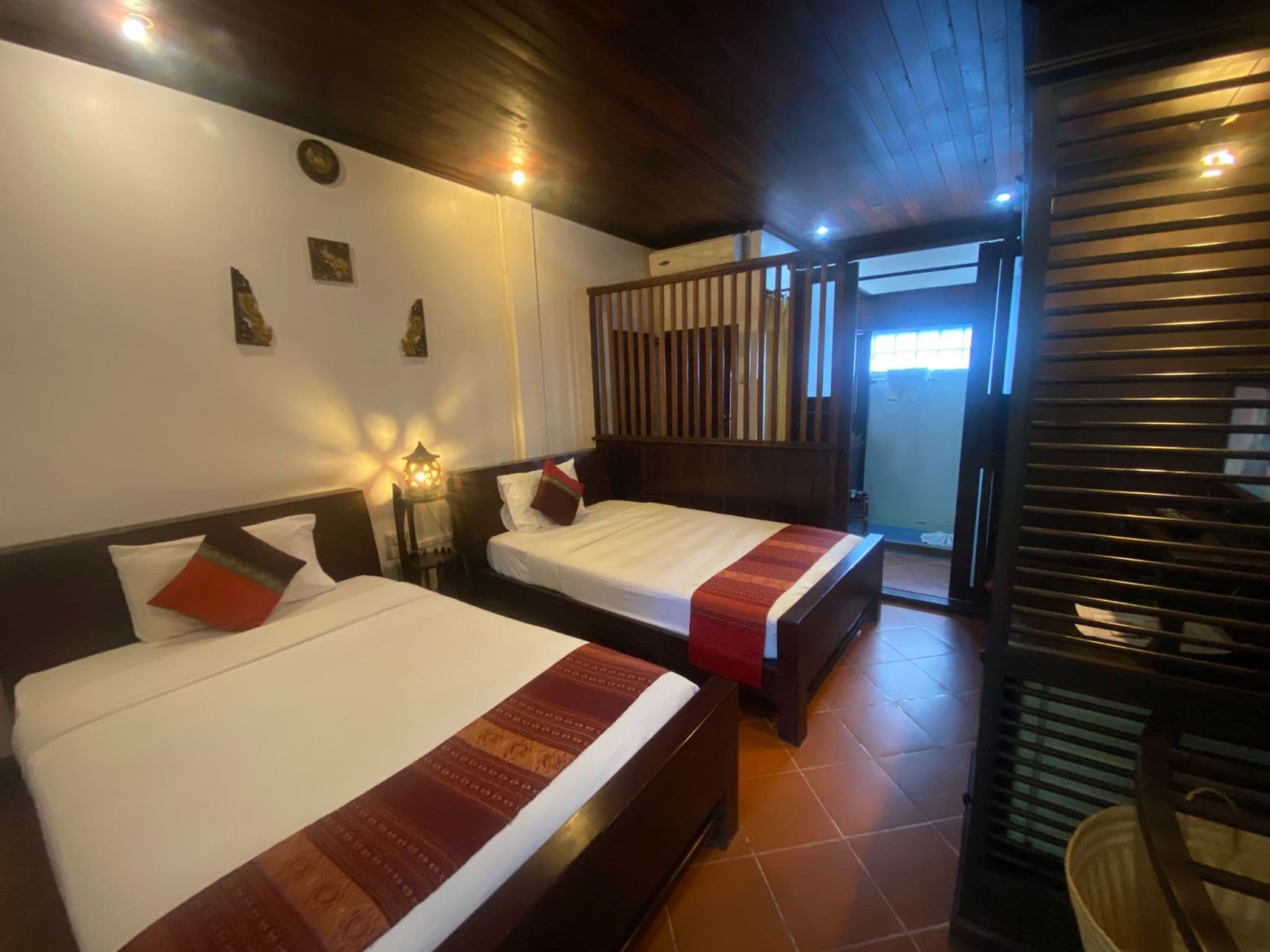 Quarto de hotel com duas camas, uma mesa de cabeceira com abajur, cabeceira de madeira e vista para o exterior.