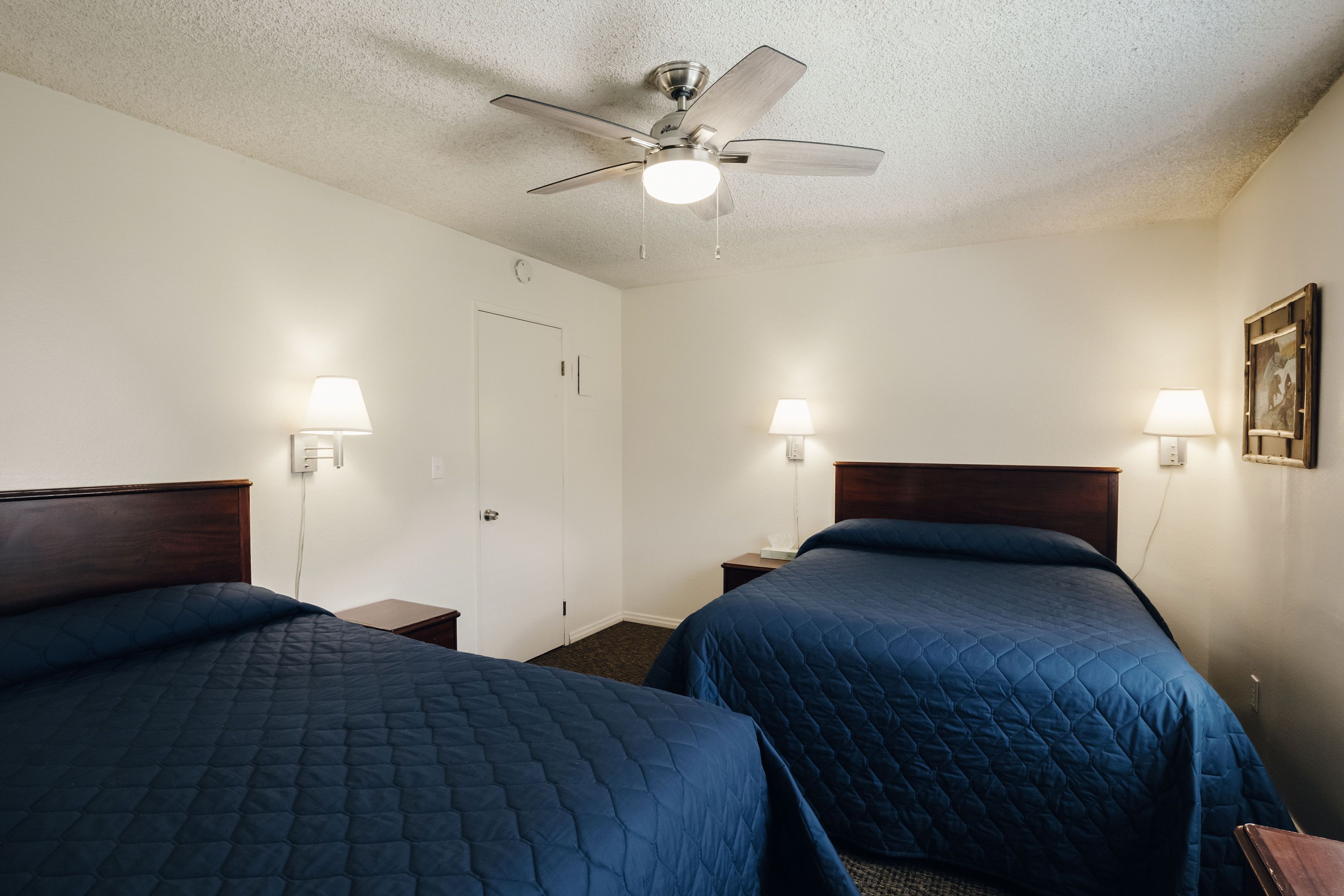 Quarto de hotel com duas camas, ventoinha no teto, luminárias de cabeceira e uma porta.