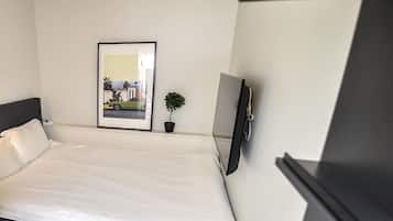 Uma cama de solteiro com colchão branco e cabeceira cinza, um pequeno vaso com planta, um quadro na parede e uma televisão fixada na parede.