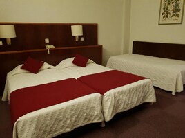 Quarto de hotel com duas camas, cada uma com colcha branca e almofadas vermelhas. Há luminárias fixadas na parede, de cada lado da cabeceira.