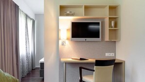 Quarto moderno com piso de madeira, sofá cinza com almofada verde, escrivaninha com cadeira, televisão de tela plana e abajur.