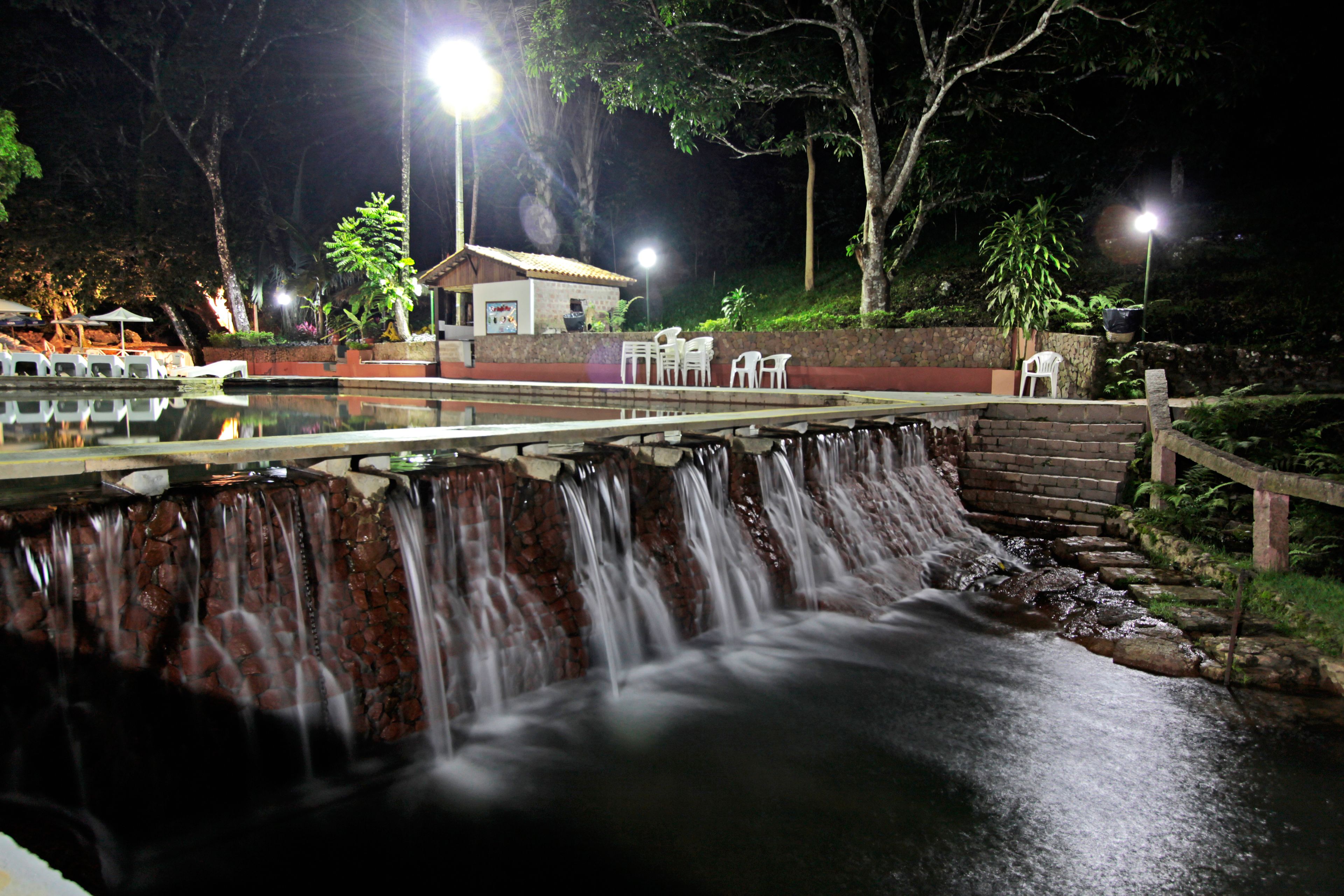 Uma cascata iluminada à noite com uma ponte de pedra, área de estar e um pequeno edifício com telhado em azulejos.