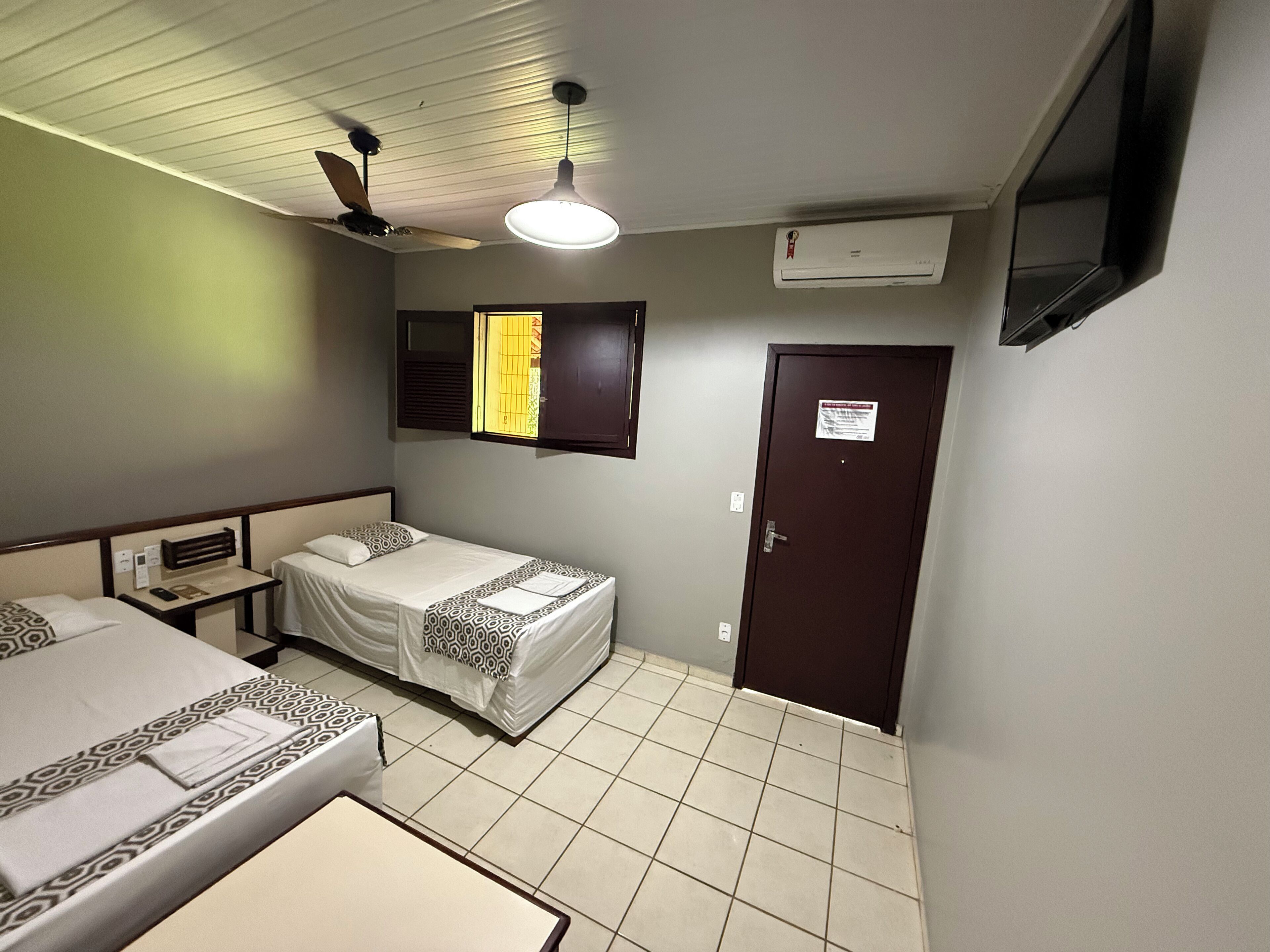 Quarto de hotel pequeno com duas camas, uma escrivaninha, ventoinha no teto, aparelho de ar condicionado e uma porta com uma placa.
