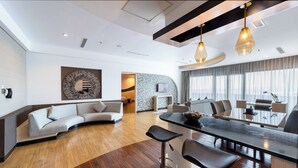 Quarto de hotel moderno com sofá curvo, área de jantar com mesa de vidro e uma janela grande com cortinas.