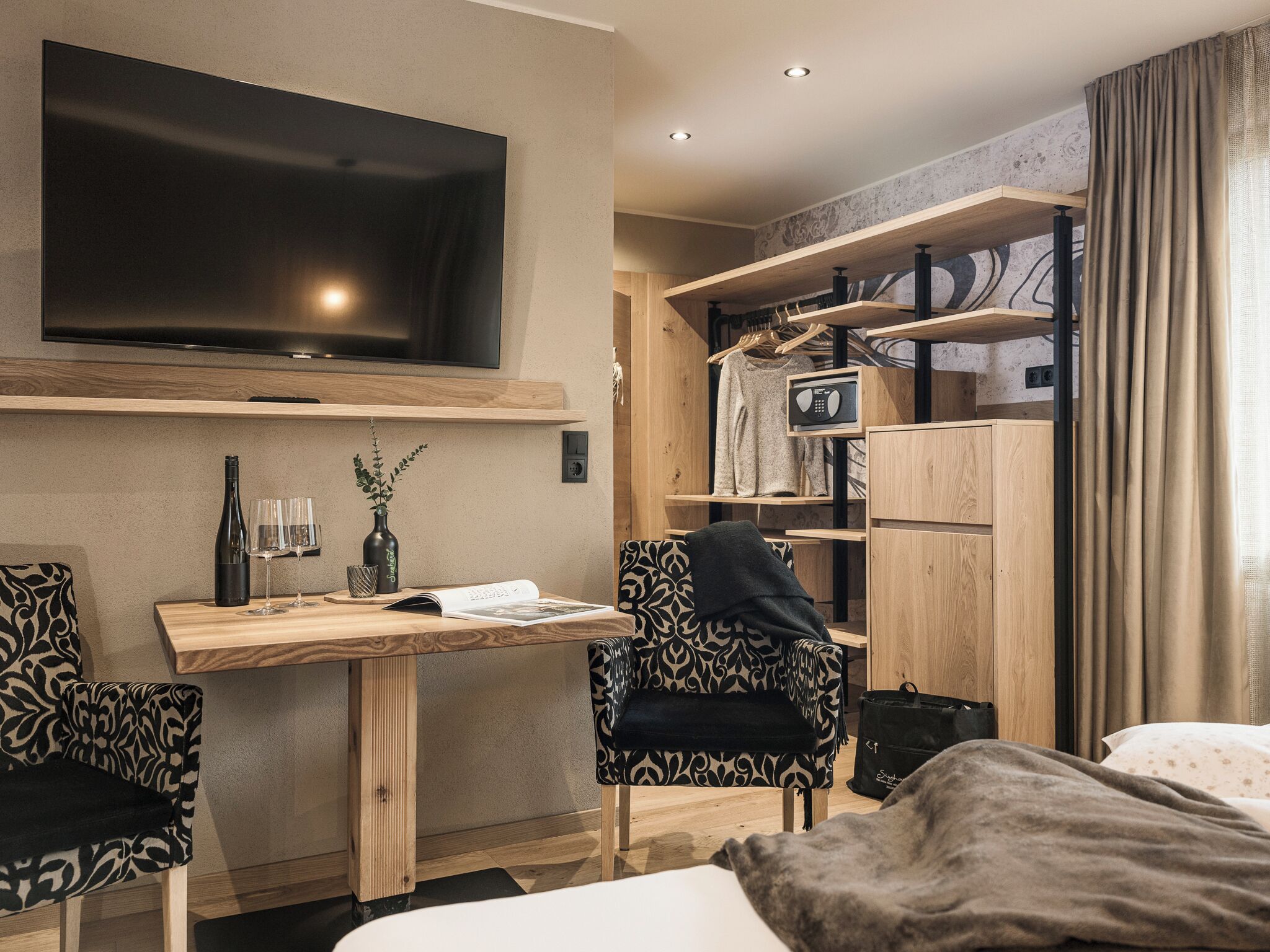 Quarto de hotel moderno com televisão de tela plana, escrivaninha de madeira, duas cadeiras estampadas, cama com cobertor cinza e nichos com roupas e um rádio.