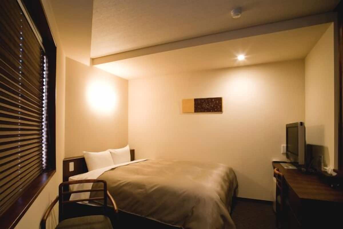 Quarto de hotel com cama, escrivaninha, cadeira, televisão e janela com persianas.