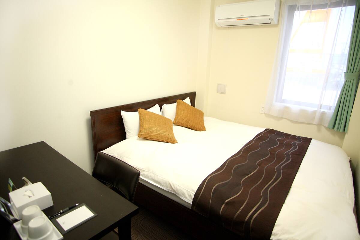 Quarto de hotel com cama, escrivaninha e cadeira, janela com cortinas e ar condicionado na parede.