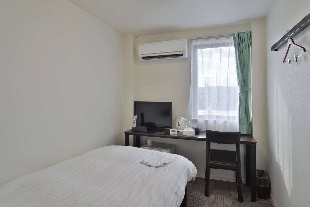 Quarto de hotel com cama, escrivaninha com computador, cadeira, ar condicionado e janela com cortinas.