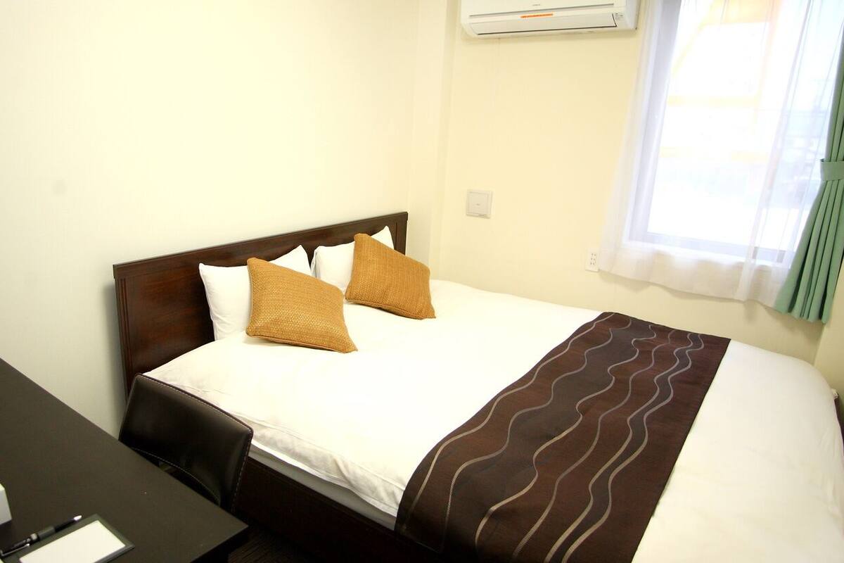 Quarto de hotel com cama, escrivaninha com cadeira, janela com cortinas e aparelho de ar condicionado.