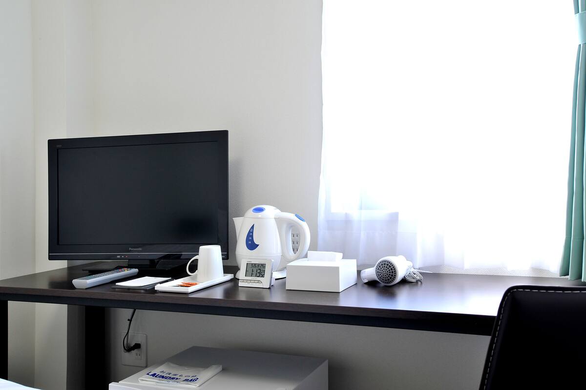 Escrivaninha de hotel com televisão, controles remotos, cafeteira, relógio, caixa de lenços, secador de cabelo e embalagem de detergente para roupa.