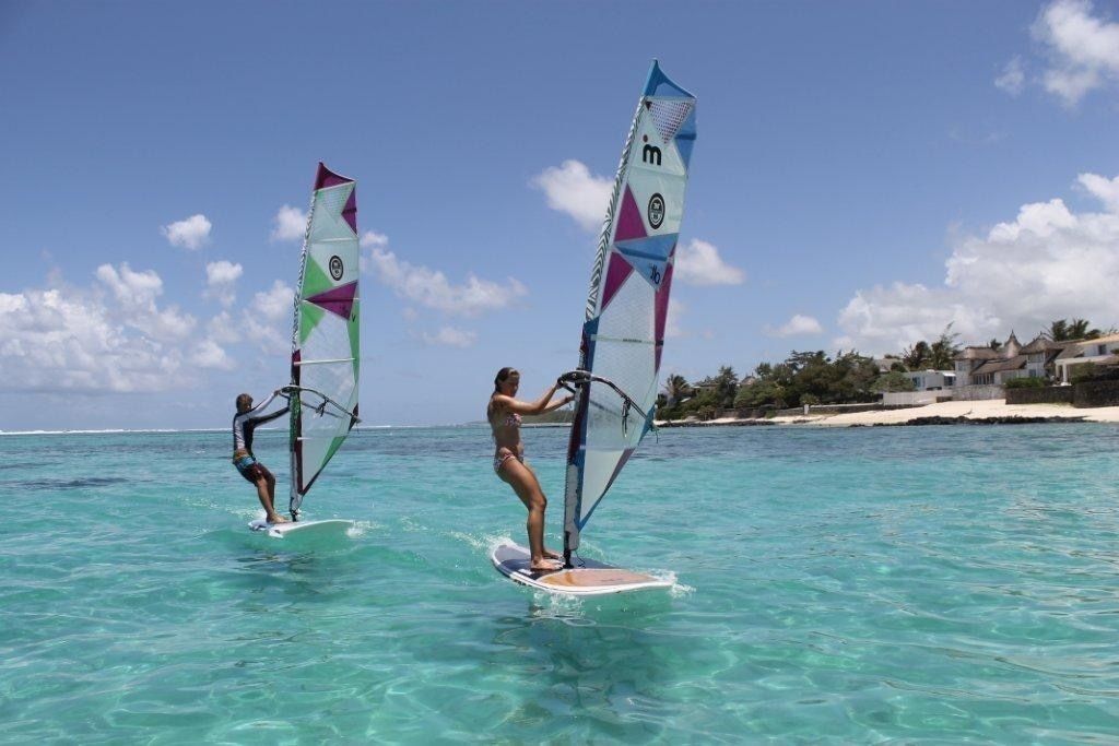 Duas pessoas praticam windsurf em águas turquesa cristalinas, com uma praia e algumas casas ao fundo.
