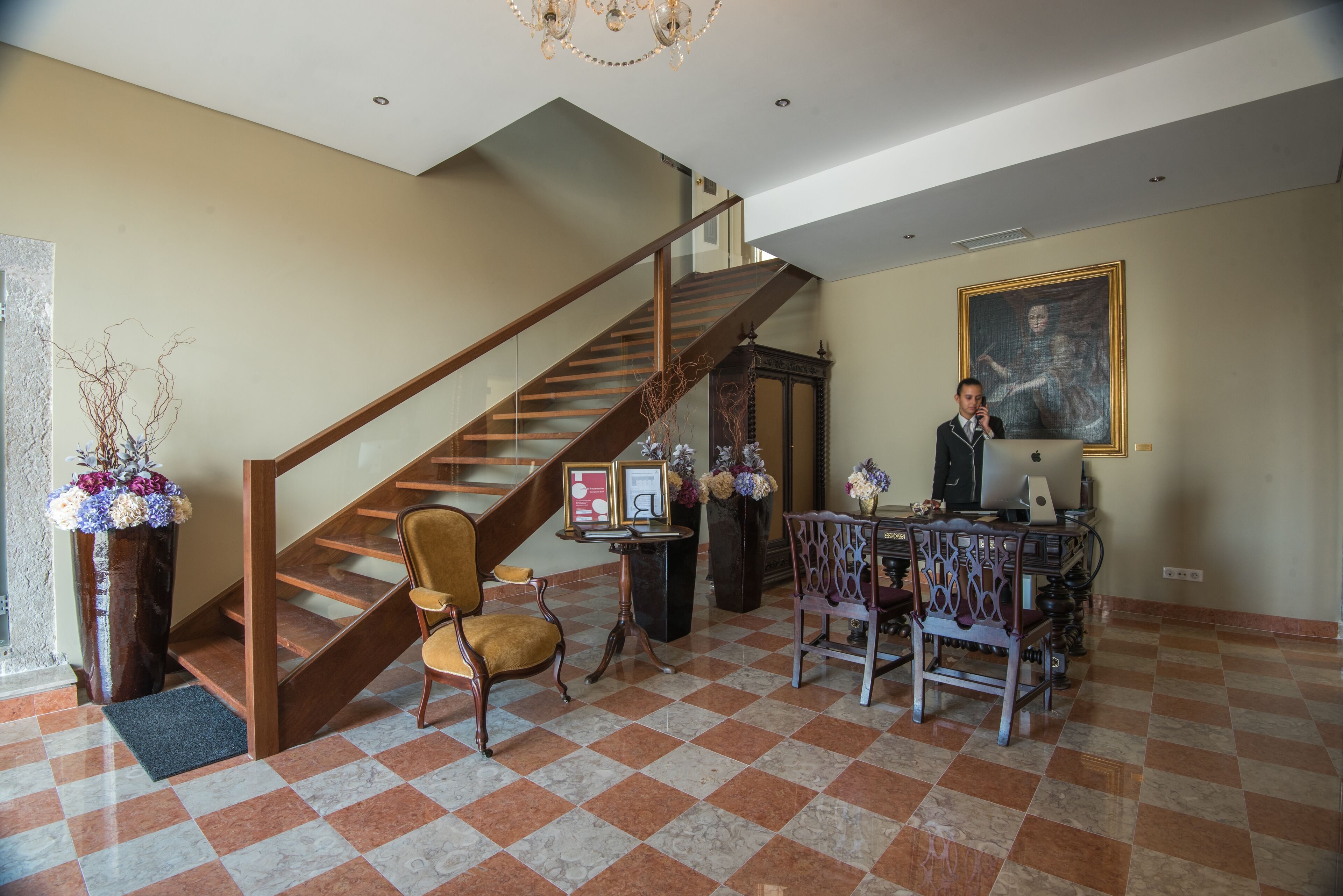 Um lobby com piso xadrez, escada de madeira, balcão de recepção e um quadro emoldurado na parede.