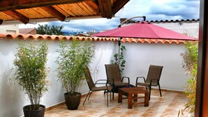 Um pátio com um guarda-sol vermelho, duas plantas em vasos e uma mesa e cadeiras de madeira.