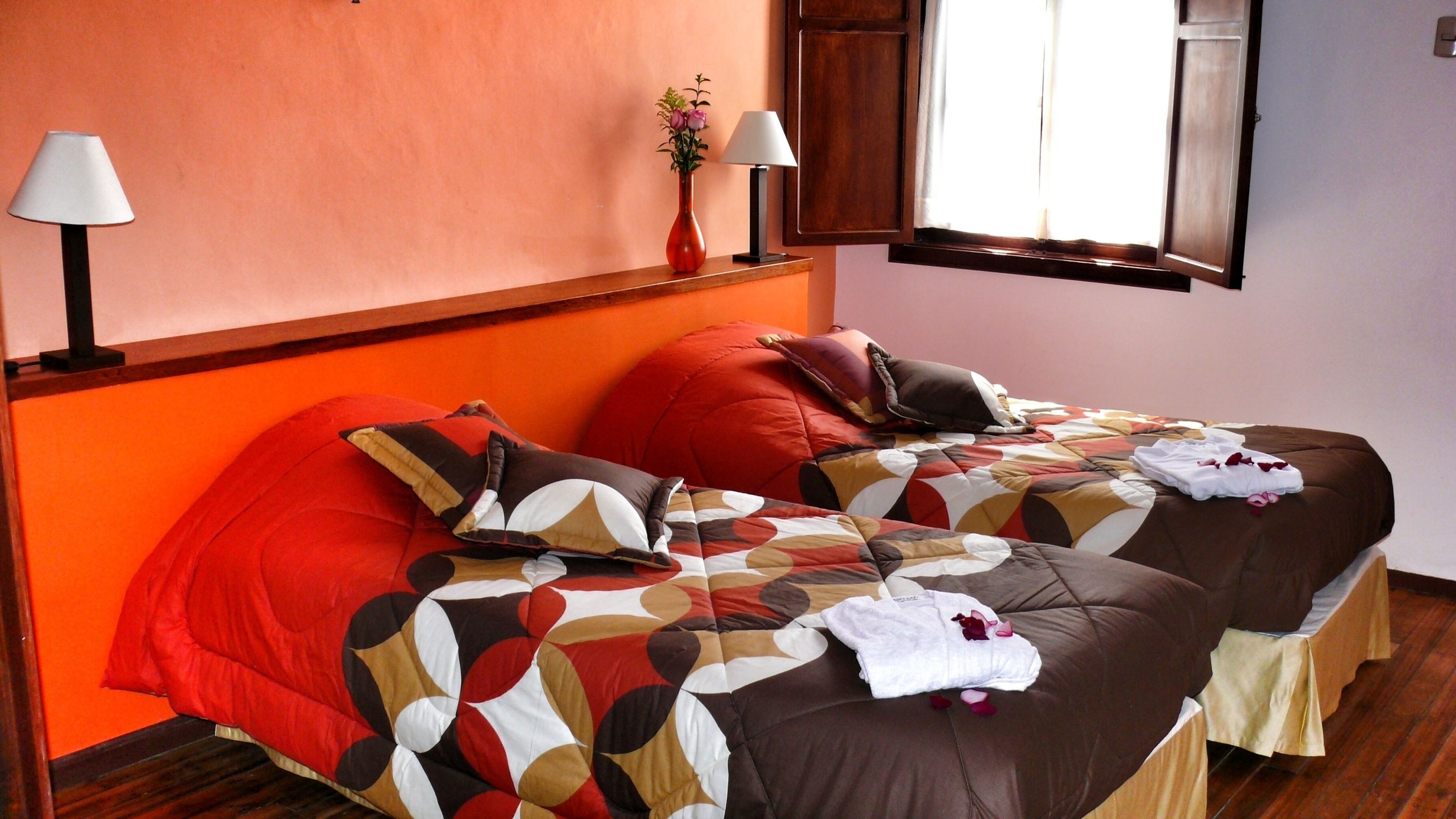 Uma cama de casal com colcha estampada, dois travesseiros, um vaso com flores e duas luminárias de cabeceira.