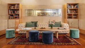 Uma sala de estar com um sofá bege, dois pufes azuis, uma mesa de centro e um tapete. Existem duas estantes de madeira de cada lado do sofá, cheias de livros e objetos decorativos.