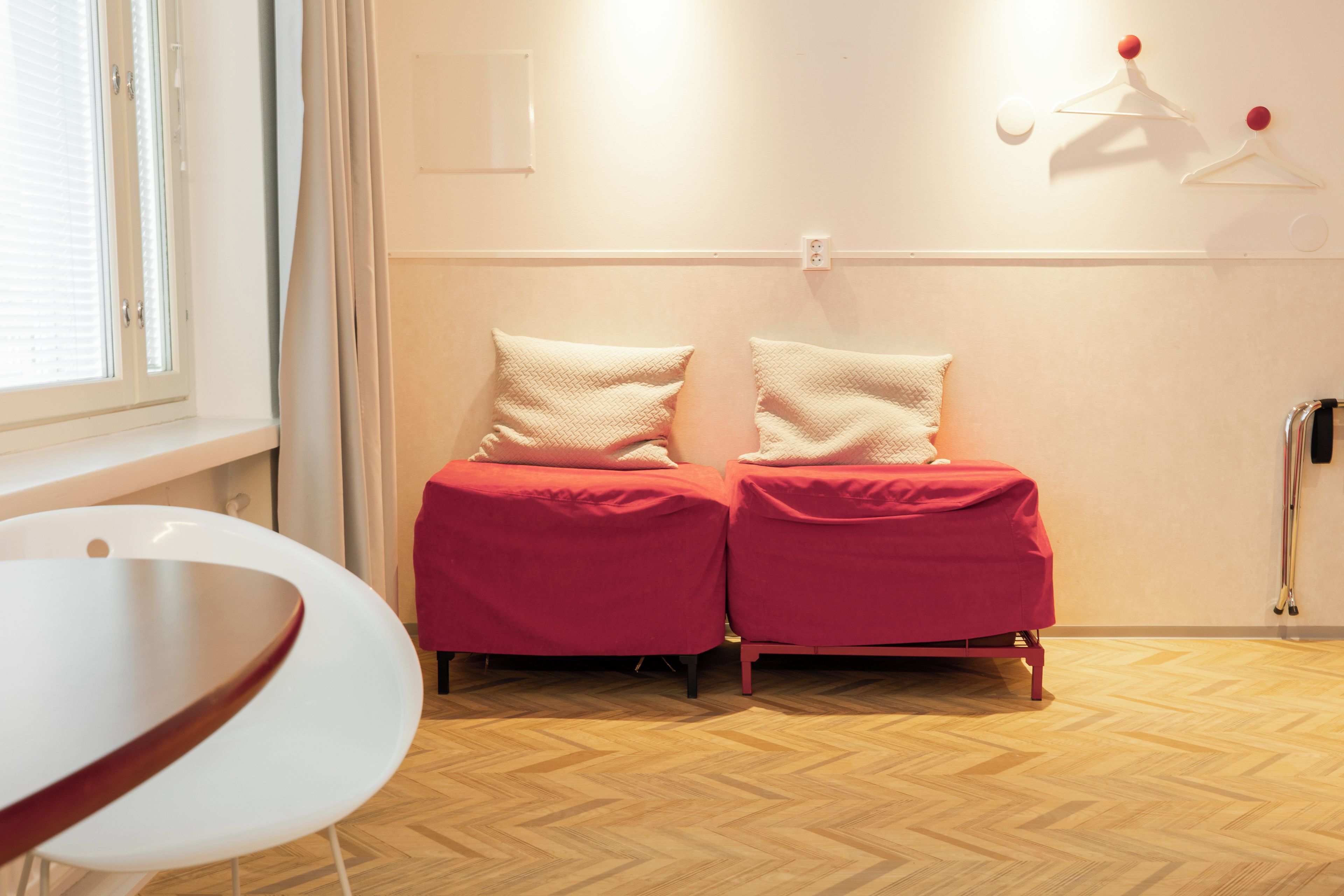 Quarto com piso de madeira em padrão herringbone, uma cadeira branca e duas camas com cobertores vermelhos.
