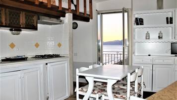 Cozinha com armários brancos, fogão e mesa de jantar com cadeiras. Vê-se o mar através de uma porta de vidro.